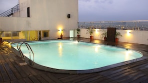 Piscina no terraço com deck de madeira, plantas em vasos e vista para a cidade ao entardecer.