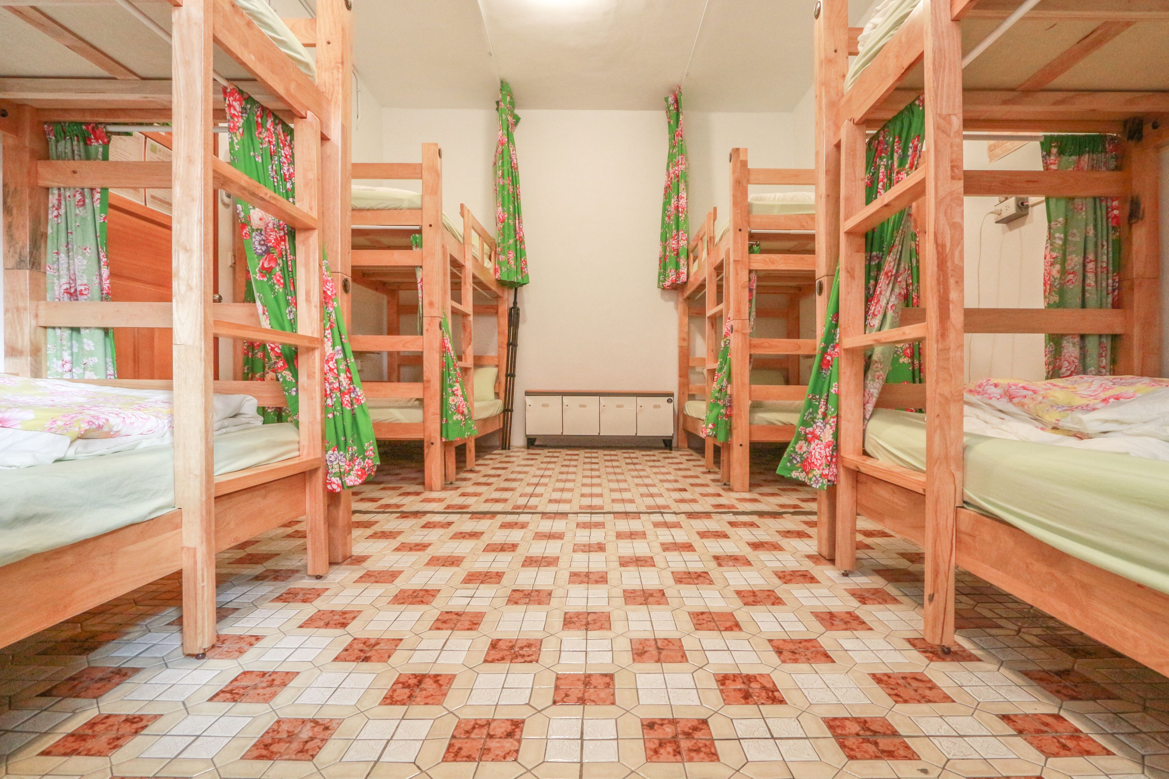 Um quarto com quatro beliches de madeira dispostos em linha, cada um com cortina florida verde. O chão tem um design de azulejos estampados.