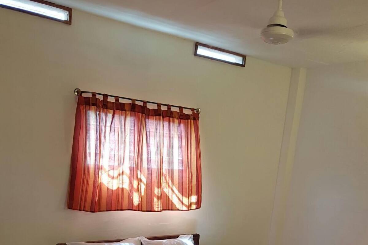 Uma cama de solteiro com cabeceira marrom, uma cadeira vermelha, uma mesinha e um ventilador de teto num quarto com paredes brancas e chão castanho.