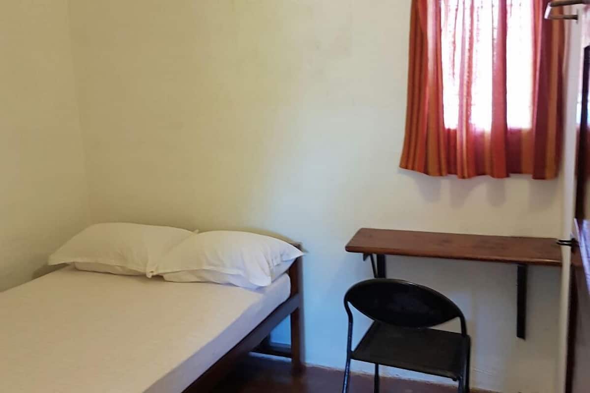 Quarto pequeno com uma cama de solteiro, ventilador de teto, janela com cortinas vermelhas, escrivaninha de madeira e uma cadeira preta.