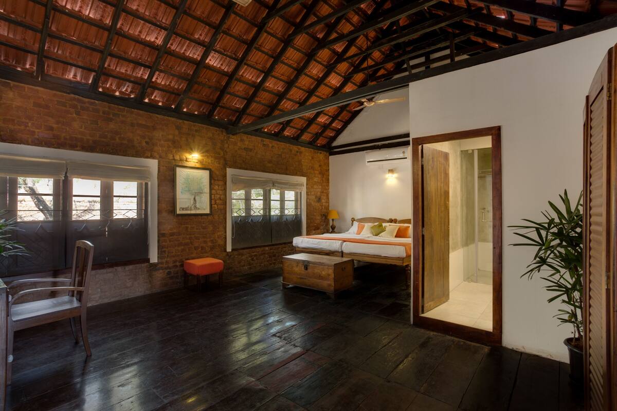 Quarto espaçoso com uma cama grande, um banco de madeira e uma cadeira. Há uma janela com vista para o exterior e uma porta que dá para o banheiro.