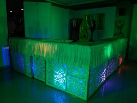 Zona de bar com um painel decorativo iluminado em verde e uma mesa com copos.