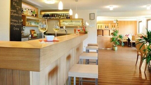 Uma área de bar com balcões e banquetas de madeira, um espaço de estar com mesas e cadeiras, e prateleiras com garrafas.