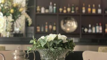 Uma mesa com jogos de chá de prata e um vaso de flores brancas, com uma estante escura ao fundo, cheia de garrafas.