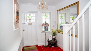 Um corredor com porta branca, tapete vermelho, lustre, espelho e uma pequena mesa com um vaso.