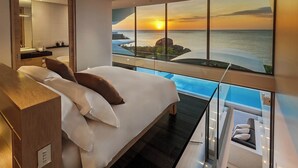 Um quarto com uma cama grande, um banheiro com pia e vista para o oceano ao pôr do sol.