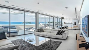 Sala de estar moderna com uma ampla parede de vidro que oferece vista para o oceano e uma varanda com mobiliário externo.