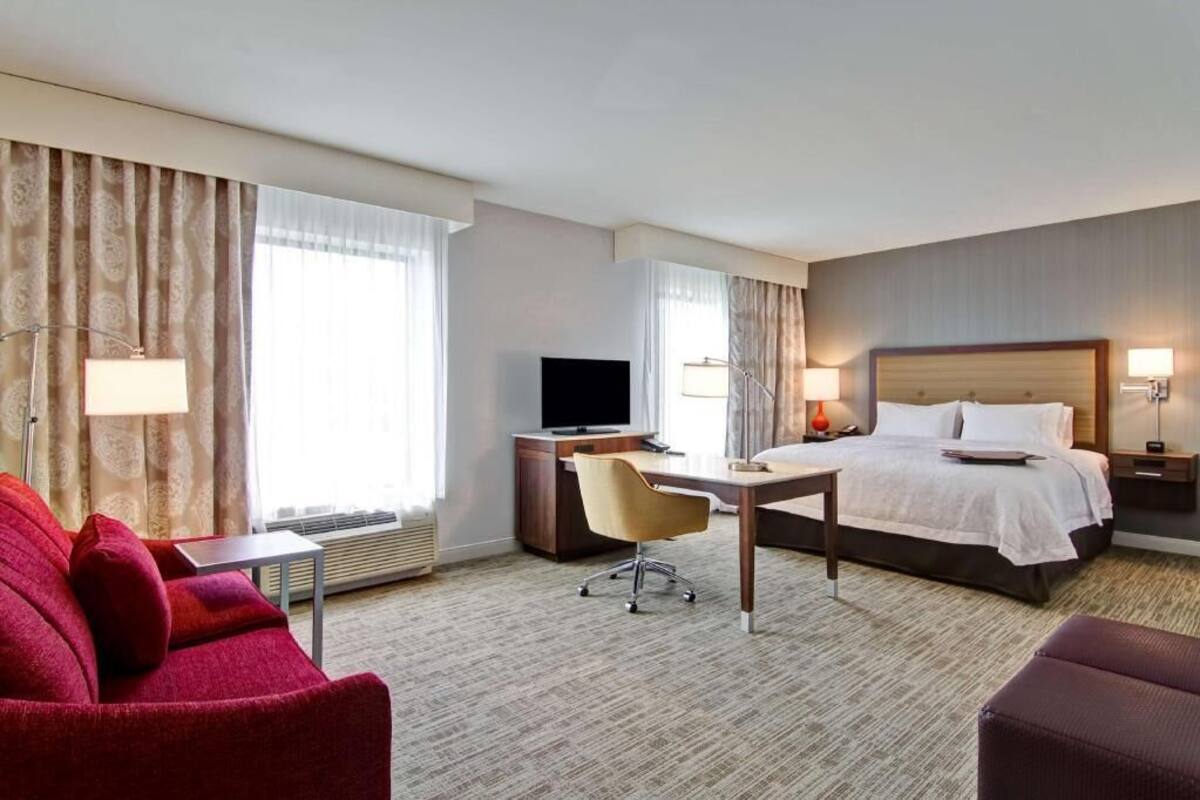 Quarto de hotel com cama, escrivaninha, televisão e duas cadeiras.