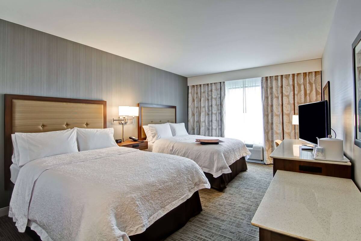 Quarto de hotel com duas camas, uma escrivaninha, televisão e janela com cortinas.
