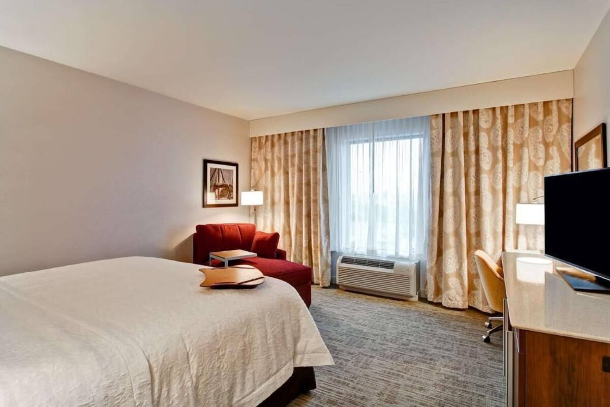 Quarto de hotel com uma cama grande, uma escrivaninha com televisão, uma cadeira vermelha e uma janela com cortinas.
