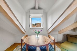 Um quarto com uma mesa de madeira redonda, cadeiras, uma janela com vista para edifícios e um pequeno vaso com flores.