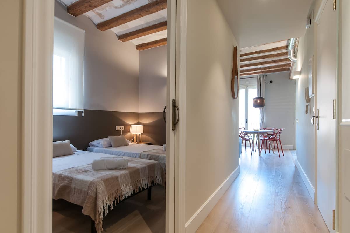 Quarto de hotel com duas camas, área de refeições com mesa e cadeiras, e corredor com vigas de madeira.
