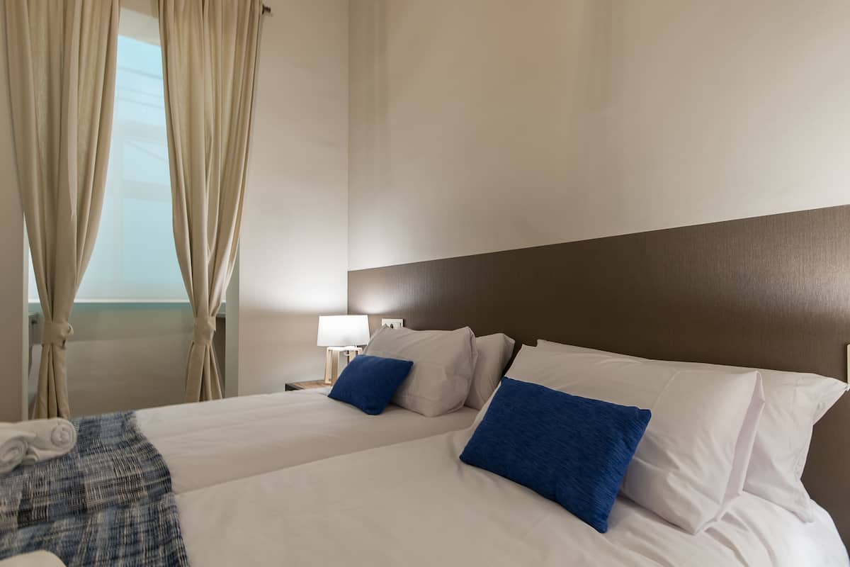 Quarto de hotel com duas camas, mesa de cabeceira, abajur e vista do céu pelas cortinas.