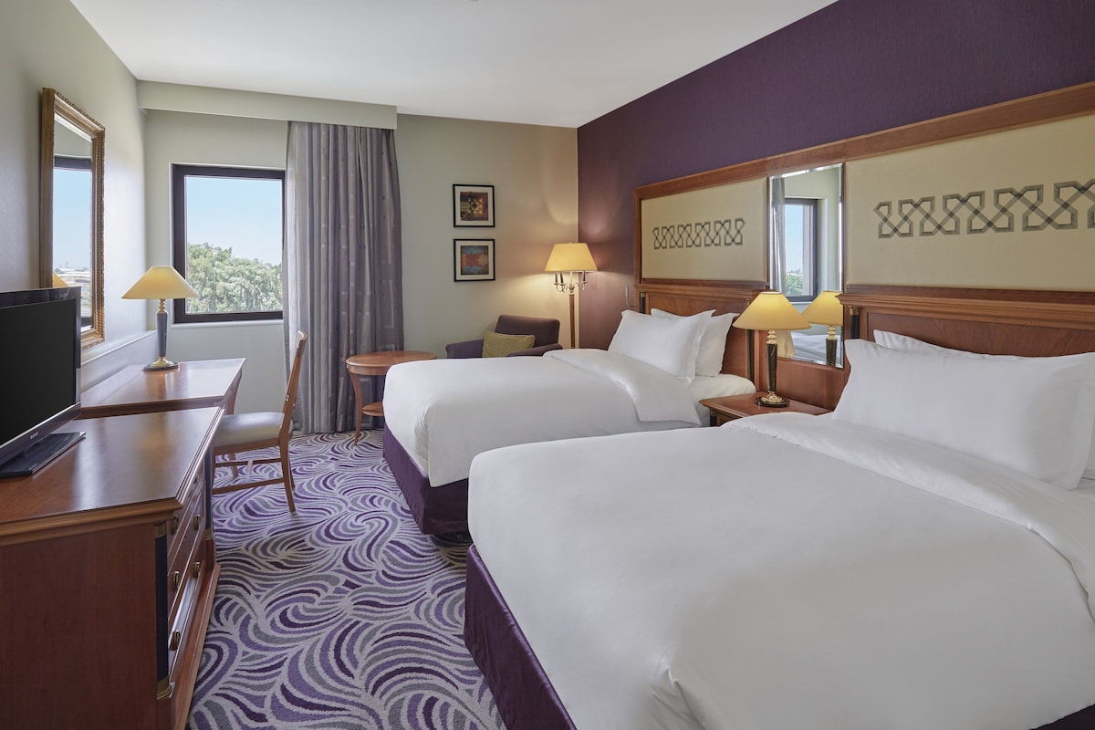 Quarto de hotel com duas camas, uma escrivaninha, uma cadeira, uma televisão e uma janela com cortinas.