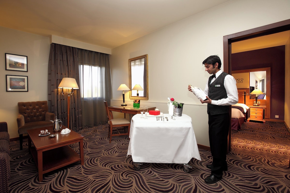 Quarto de hotel com mesa de jantar posta, sofá, poltrona, luminária e espelho.