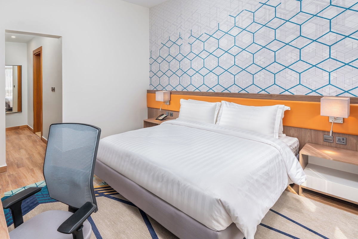Quarto de hotel com cama, escrivaninha, cadeira, mesa de cabeceira e parede estampada.