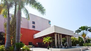Um edifício moderno com fachada vermelha, área externa coberta com colunas e uma fonte.