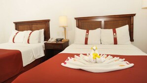 Quarto de hotel com duas camas, cada uma com cabeceira de madeira, roupa de cama branca e detalhes em vermelho. Há uma mesa de cabeceira com abajur e telefone.