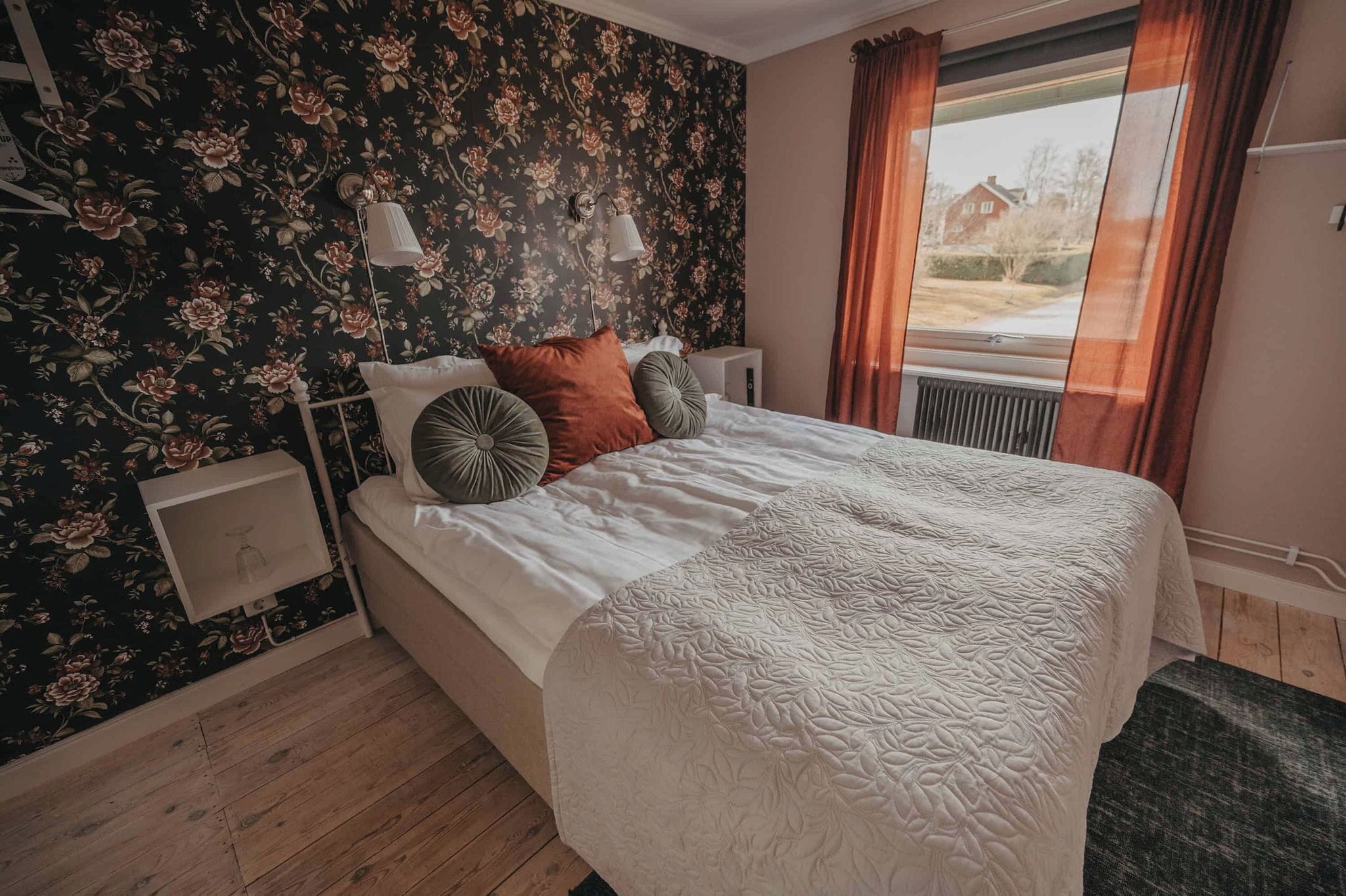 Quarto com papel de parede floral, cama branca com almofadas decorativas, mesa de cabeceira com abajur e janela com cortinas.