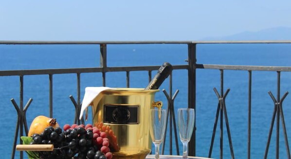 Uma mesa com um balde de champanhe, uma romã, uvas e uma taça de champanhe em uma varanda com vista para o mar.