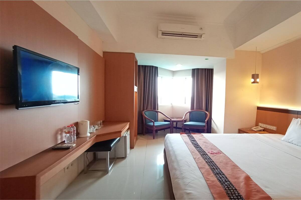 Quarto de hotel com uma cama, uma secretária com computador, duas cadeiras, uma televisão e um quadro na parede.