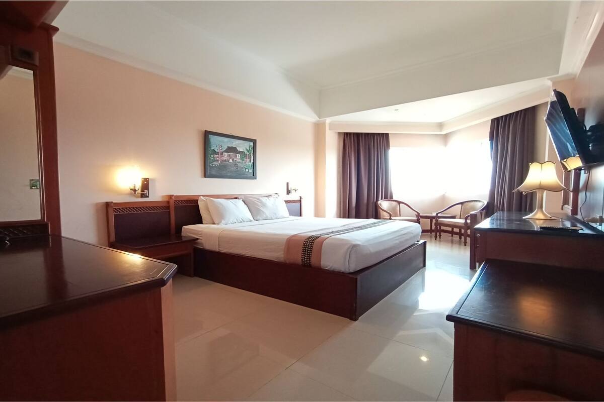 Quarto de hotel com cama, escrivaninha, cadeira, luminária e um quadro na parede.