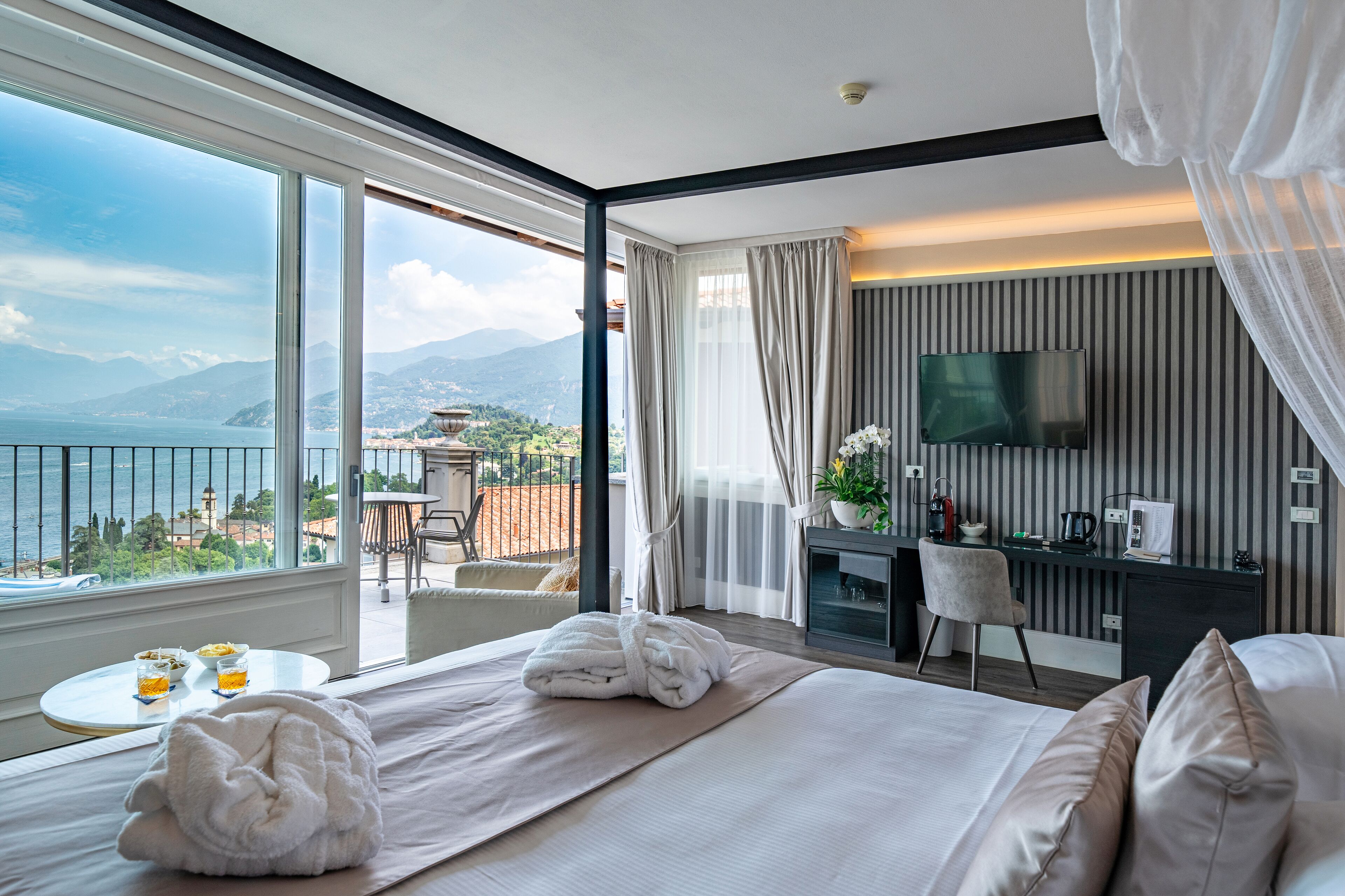 Quarto de hotel com uma janela ampla que oferece vista para o mar e montanhas, uma cama com toalhas, uma mesa com televisão e uma varanda com mesa e cadeiras.