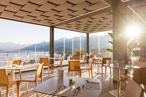 Uma sala de jantar moderna com mesas e cadeiras, janelas amplas com vista para montanhas e um corpo d'água.