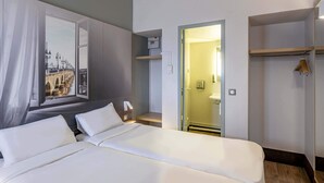 Quarto de hotel com uma cama grande, janela com vista para uma ponte, banheiro com chuveiro e prateleira de madeira com cabides.