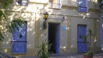 Um edifício com fachada tradicional, apresentando uma entrada central com porta e janelas com persianas azuis. Há plantas em vasos na frente da entrada.