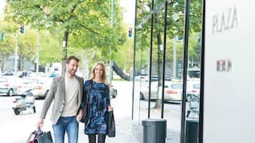 Um casal caminhando em uma calçada carregando sacolas de compras, em frente a um edifício com fachada de vidro e a palavra "PLAZA" visível.