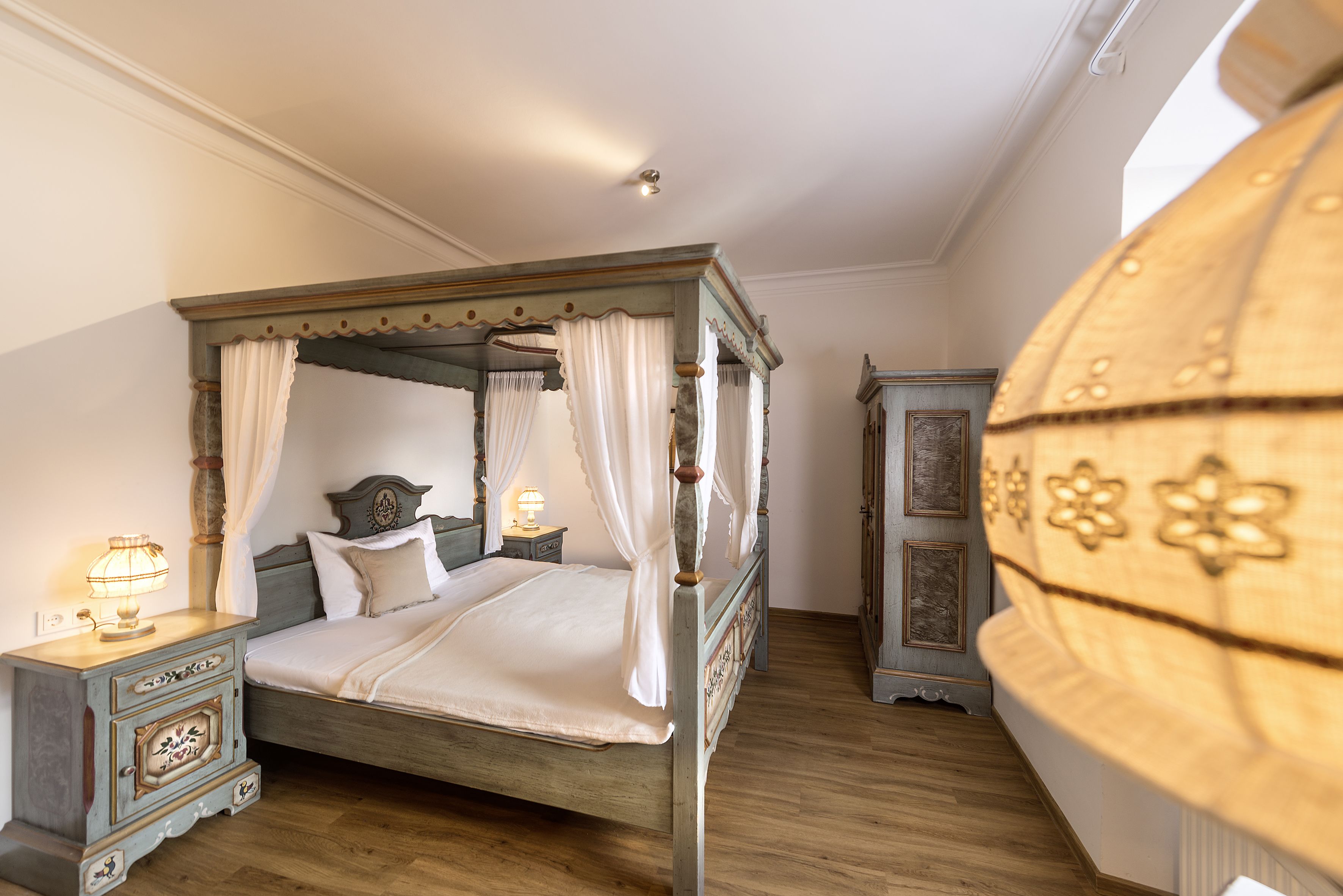 Uma cama de dossel com lençóis brancos, uma mesa de cabeceira com abajur e chão de madeira.