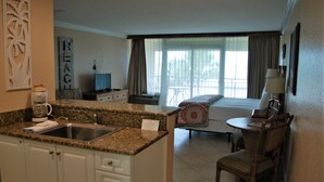 Quarto de hotel com cozinha, mesa de jantar com cadeiras, cama e varanda com cortinas.