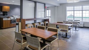 Uma sala de jantar moderna com várias mesas e cadeiras, área de cozinha com eletrodomésticos, e grandes janelas que proporcionam luz natural.