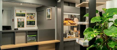 Uma área de recepção com balcão, cartazes informativos, uma planta e um vislumbre de uma cozinha.