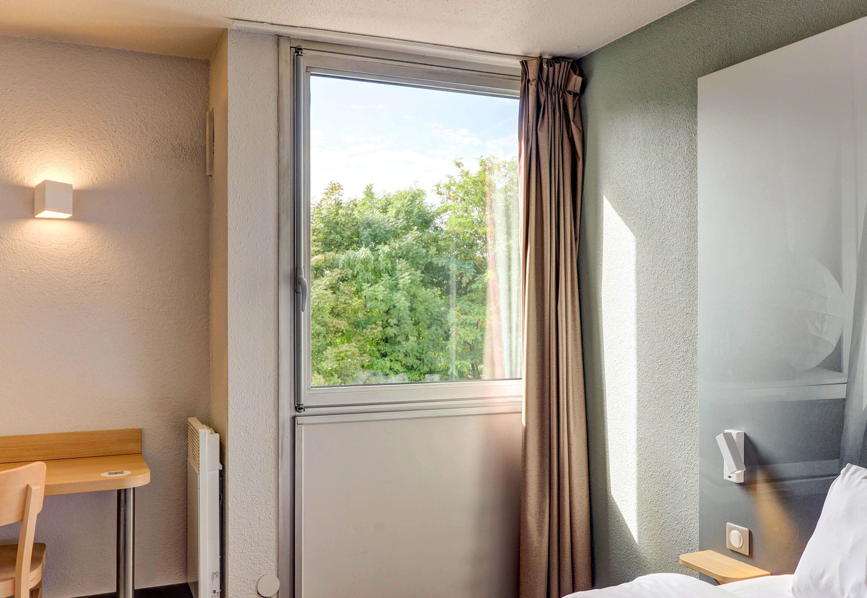 Quarto de hotel com janela, escrivaninha, cadeira, cama e mesa de cabeceira.