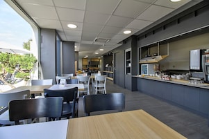 Um restaurante moderno com mistura de mesas internas e externas, área de cozinha e grandes janelas com vista para o exterior.