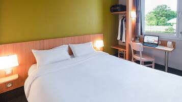Quarto de hotel com cama, escrivaninha com laptop, cadeira e janela com vista para árvores.