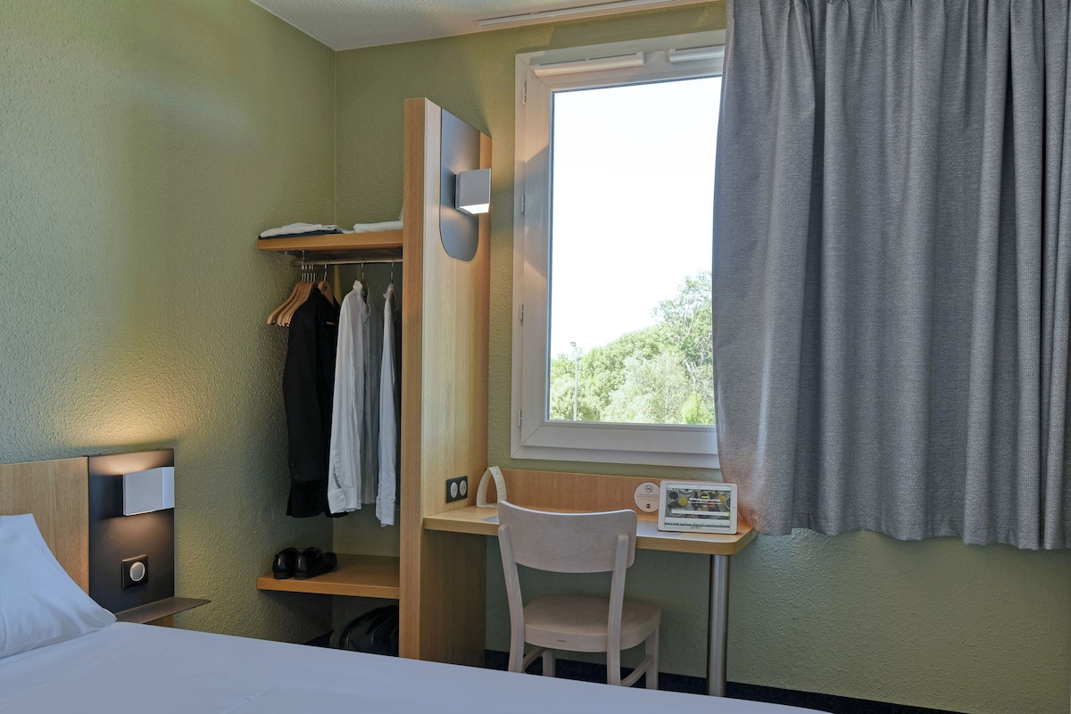 Quarto de hotel com cama, escrivaninha com cadeira, guarda-roupa e janela com cortinas.