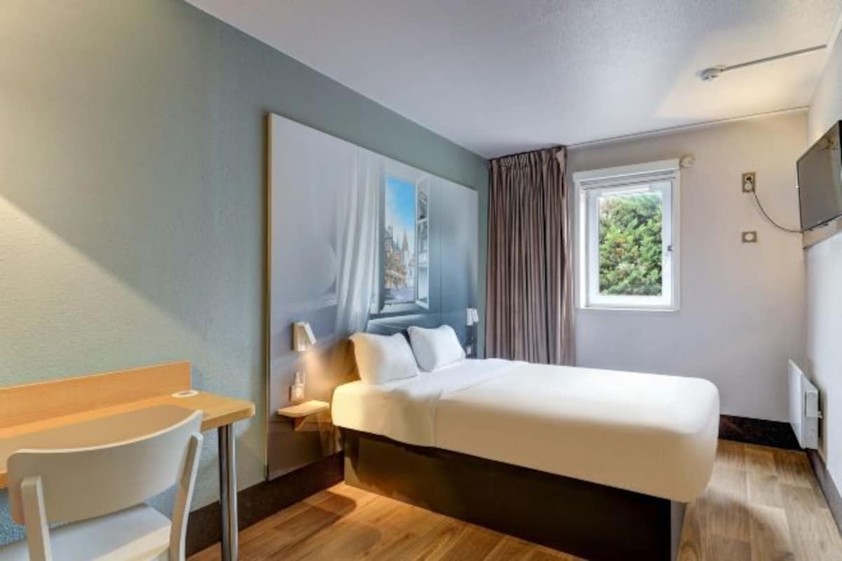 Quarto de hotel com cama, escrivaninha, cadeira, quadro na parede, janela com cortinas e televisão fixada na parede.