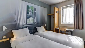 Quarto de hotel com duas camas, uma escrivaninha pequena, uma janela com cortinas e uma luminária na parede.