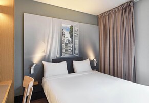 Quarto de hotel com cama, duas mesinhas de cabeceira, uma luminária de parede e uma janela grande com cortinas.