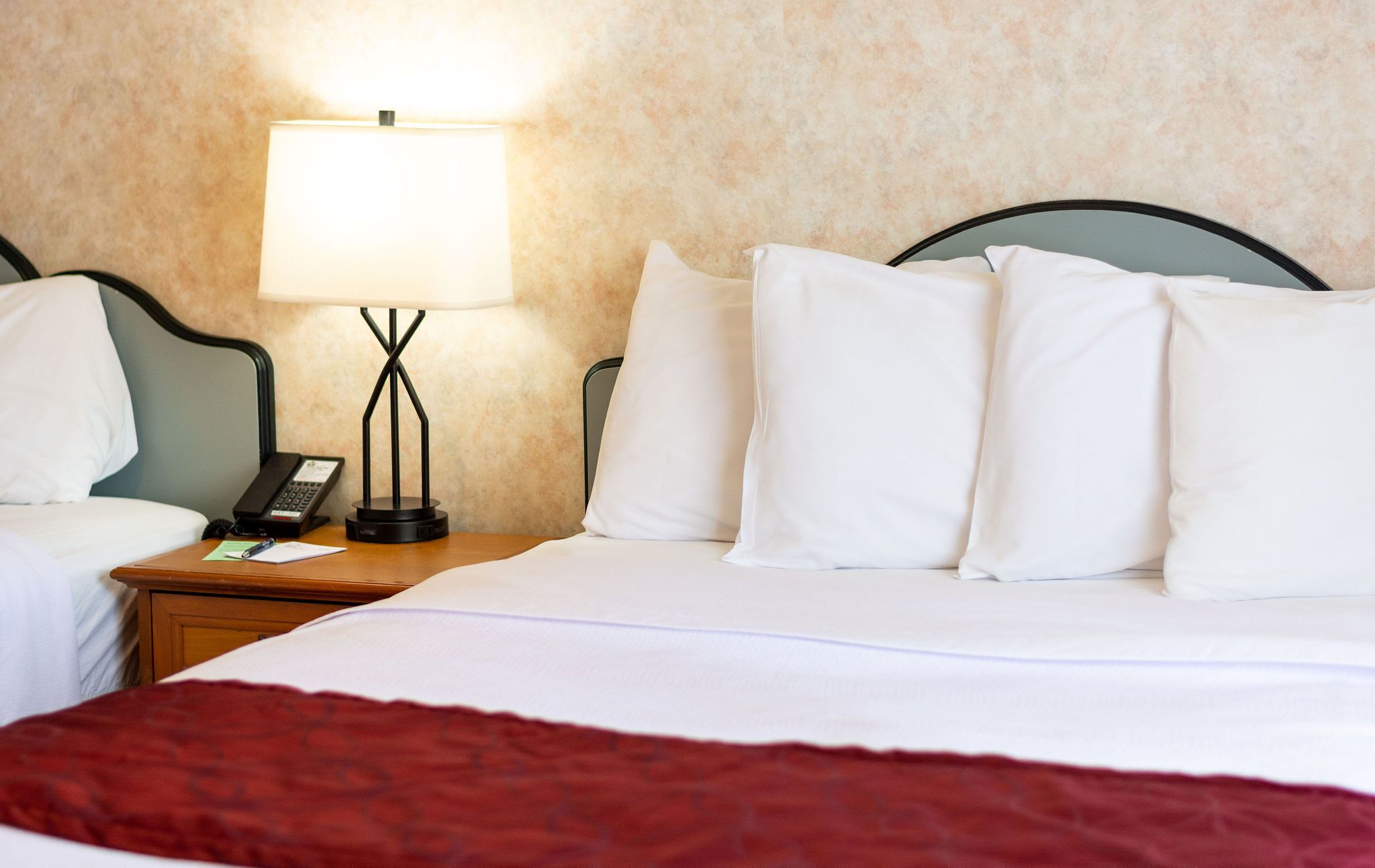 Quarto de hotel com cama, almofadas, mesa de cabeceira com abajur e telefone.