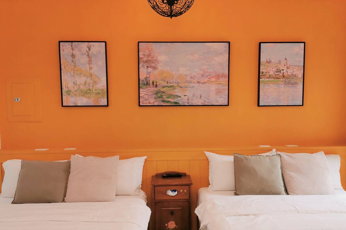 Quarto de hotel com duas camas, uma mesa de cabeceira e três quadros na parede.