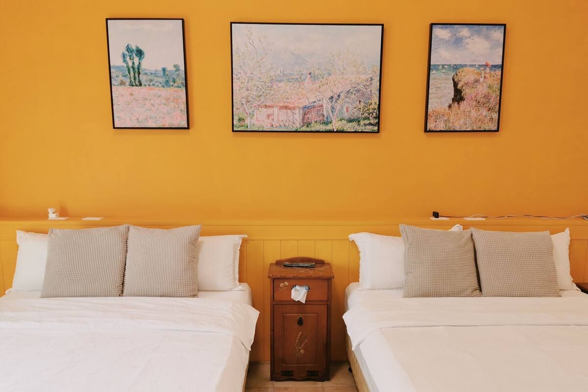 Quarto de hotel com duas camas, uma mesa de cabeceira de madeira e três quadros na parede.