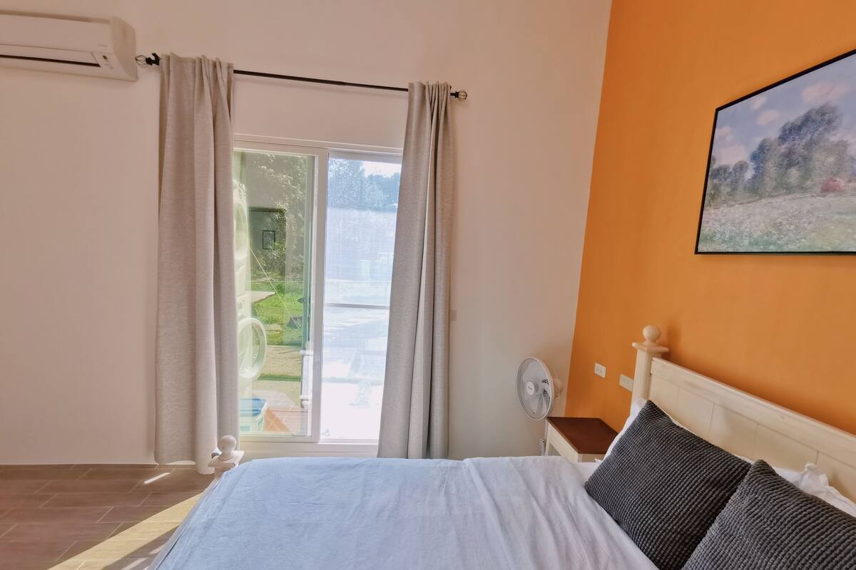 Quarto com cama, mesa de cabeceira, ventoinha na parede e janela com cortinas.