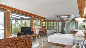 Quarto moderno com mesa de centro de vidro, cama, TV e área de refeições com banquetas.