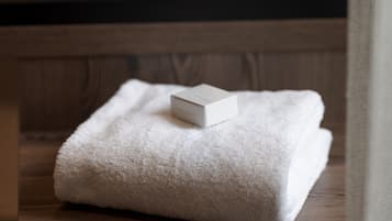 Uma toalha branca dobrada cuidadosamente sobre uma superfície de madeira, com um pequeno objeto branco em cima, e um banheiro com banheira e box de vidro ao fundo.