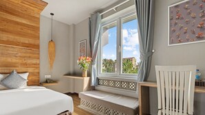 Quarto de hotel com cabeceira de madeira, cama grande com roupa de cama branca, escrivaninha com cadeira, janela com vista para palmeiras e luminária pendente.