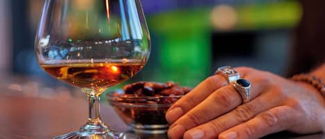 Um copo com líquido âmbar sobre uma superfície de madeira, uma pequena tigela com objetos escuros e uma mão com um anel.